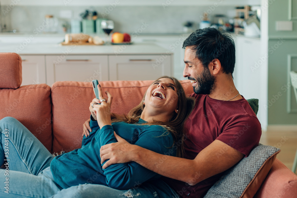 happy couple hugging using cellphone together lying on sofa at home ...