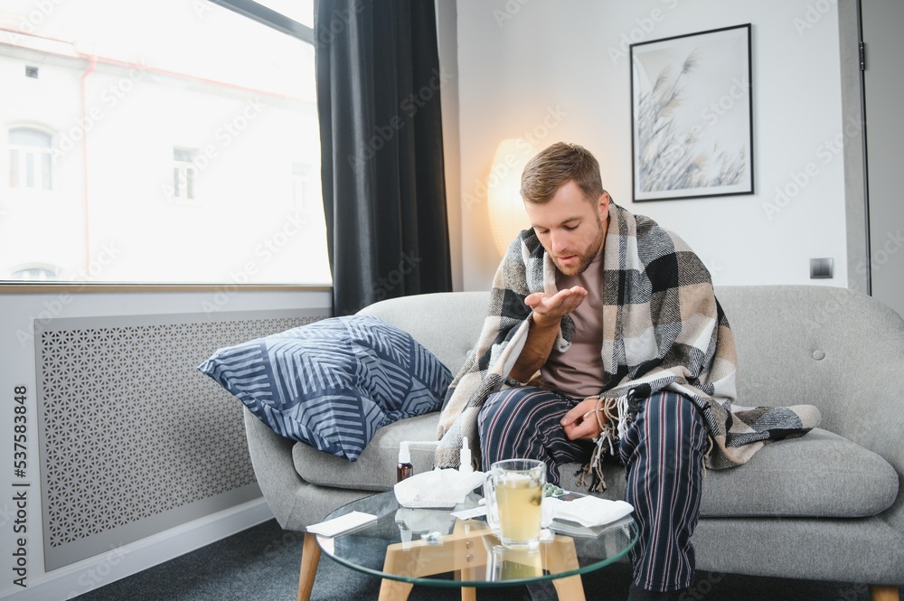 Young man suffering from cold at his home. Shot of a young man sitting ...