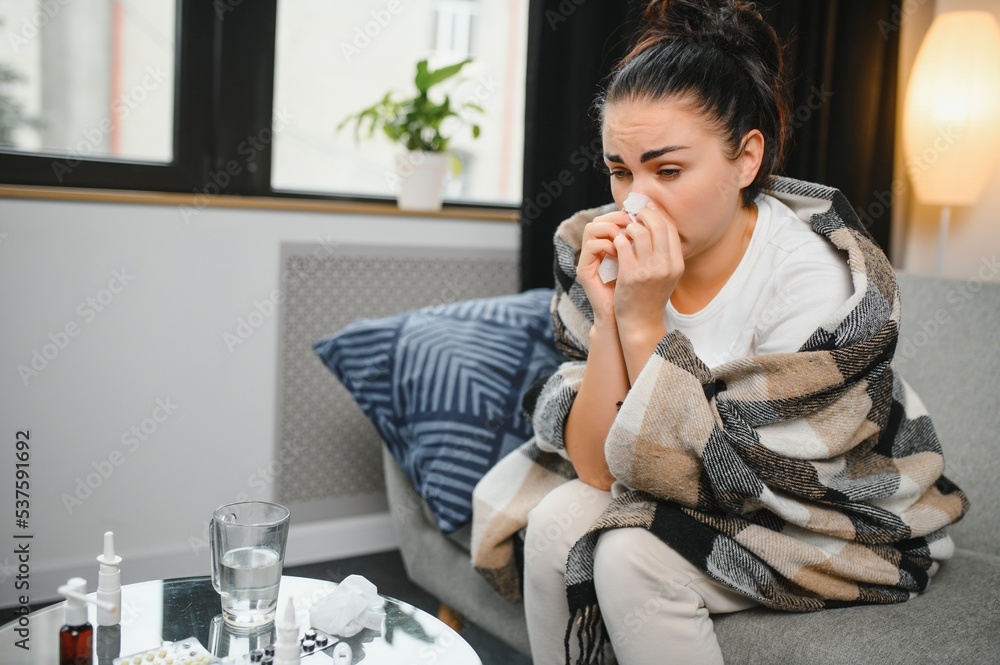 Ill young woman sit on sofa covered with blanket, freezing, blowing her ...