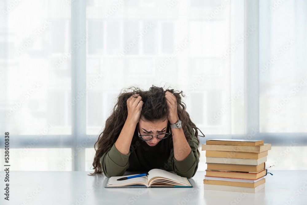 Young frustrated school girl sitting at desk with pile of books feeling ...
