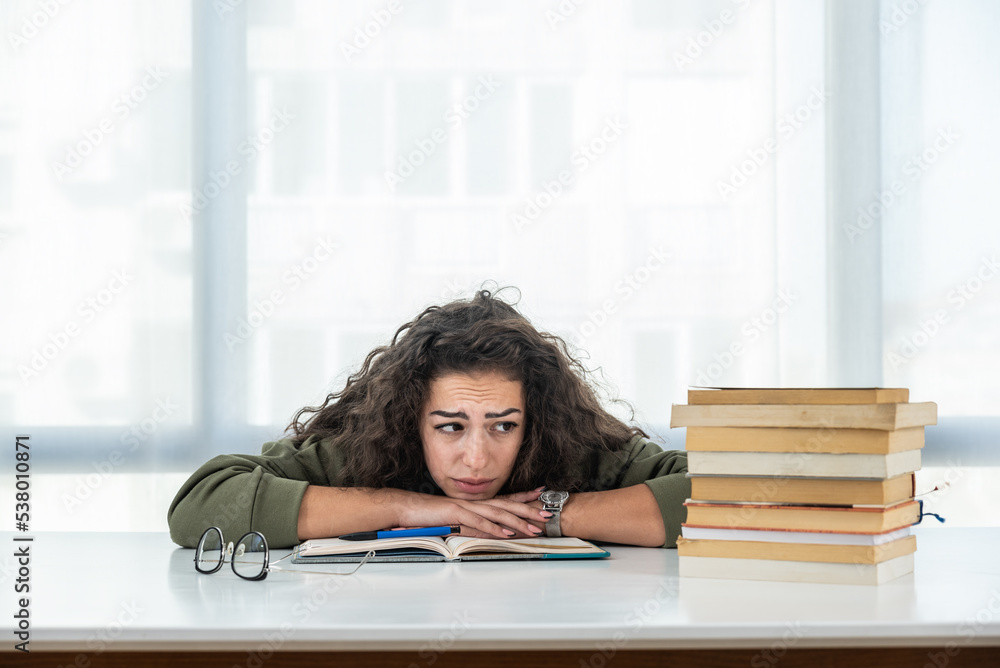 Young frustrated school girl sitting at desk with pile of books feeling ...