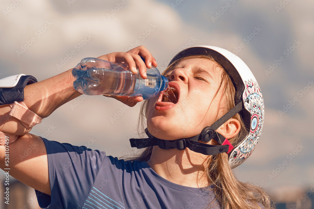 A 10-12 year old child in a protective helmet greedily drinks water from a bottle after playing ...