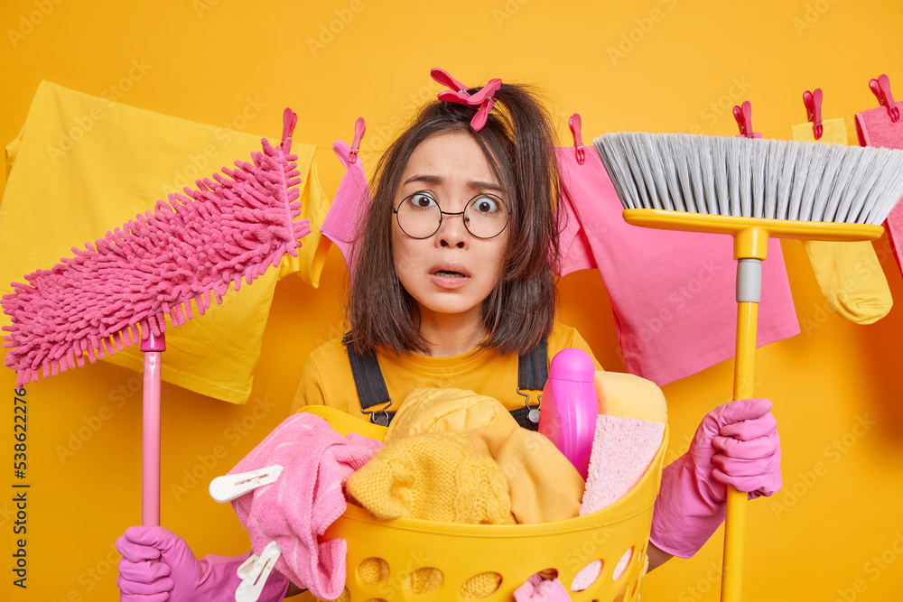 Embarrassed Shocked Asian Woman With Dark Hair Holds Cleaning Mop And Broom Stares At Camera Has
