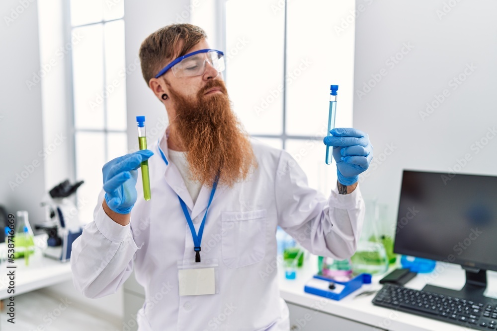 Young redhead man wearing scientist uniform holding test tubes at ...