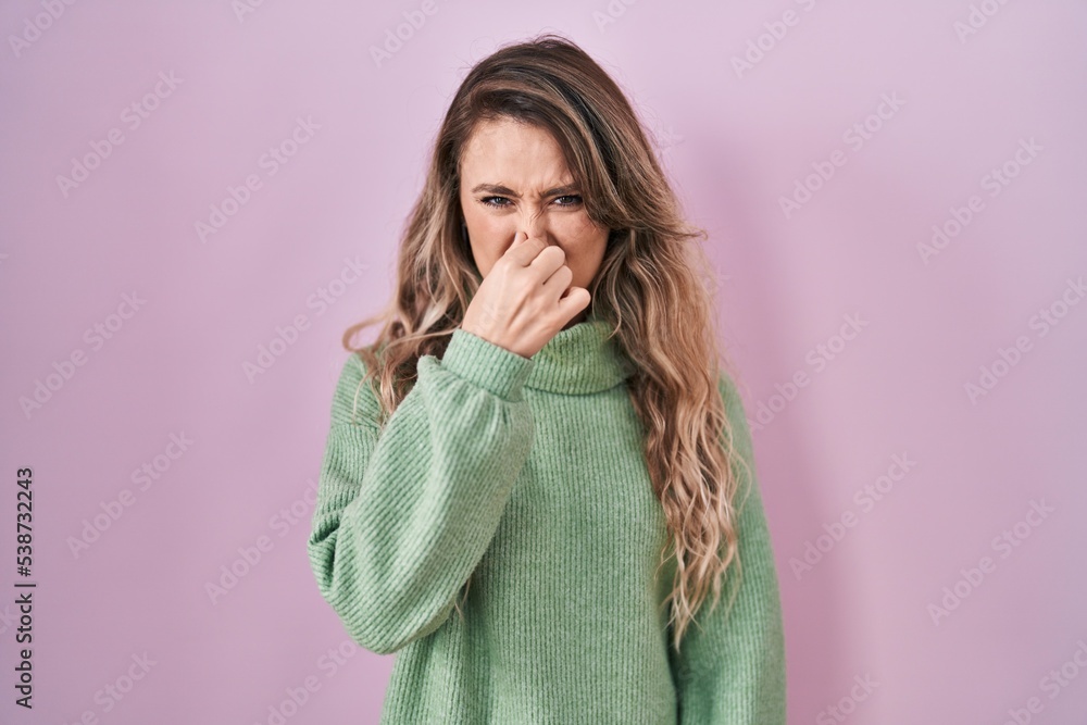 Young caucasian woman standing over pink background smelling something ...