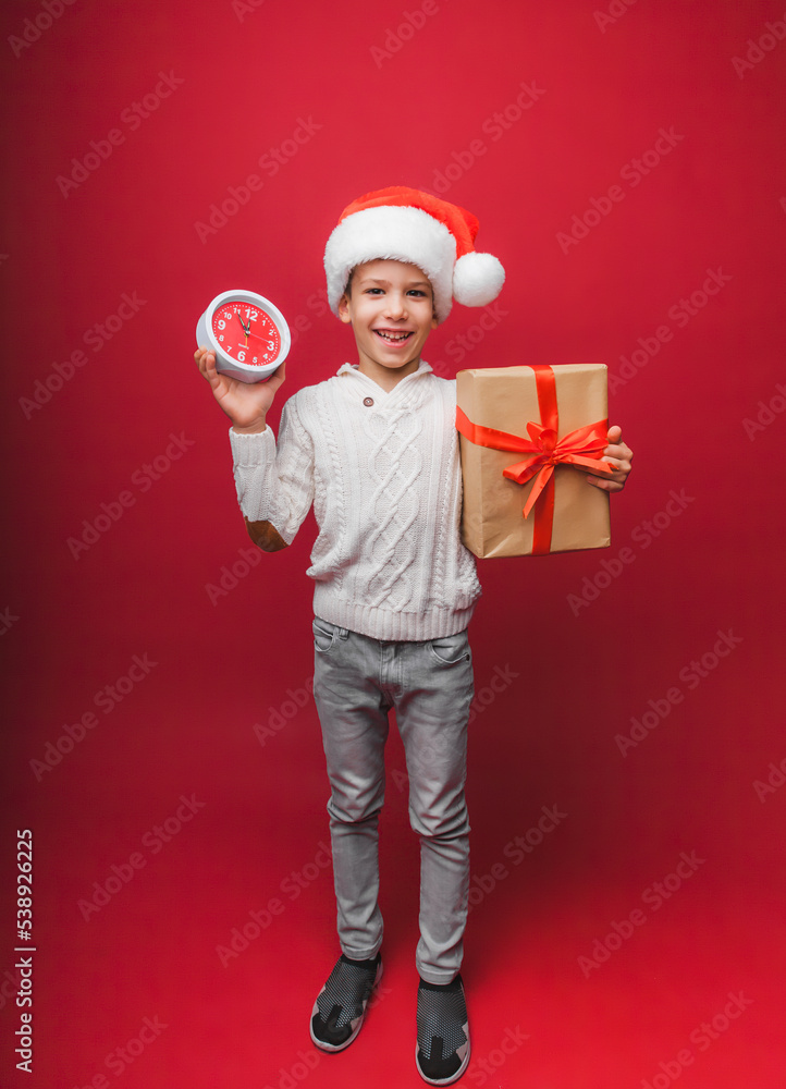 A boy in a Santa Claus hat holds a watch on a red studio background ...