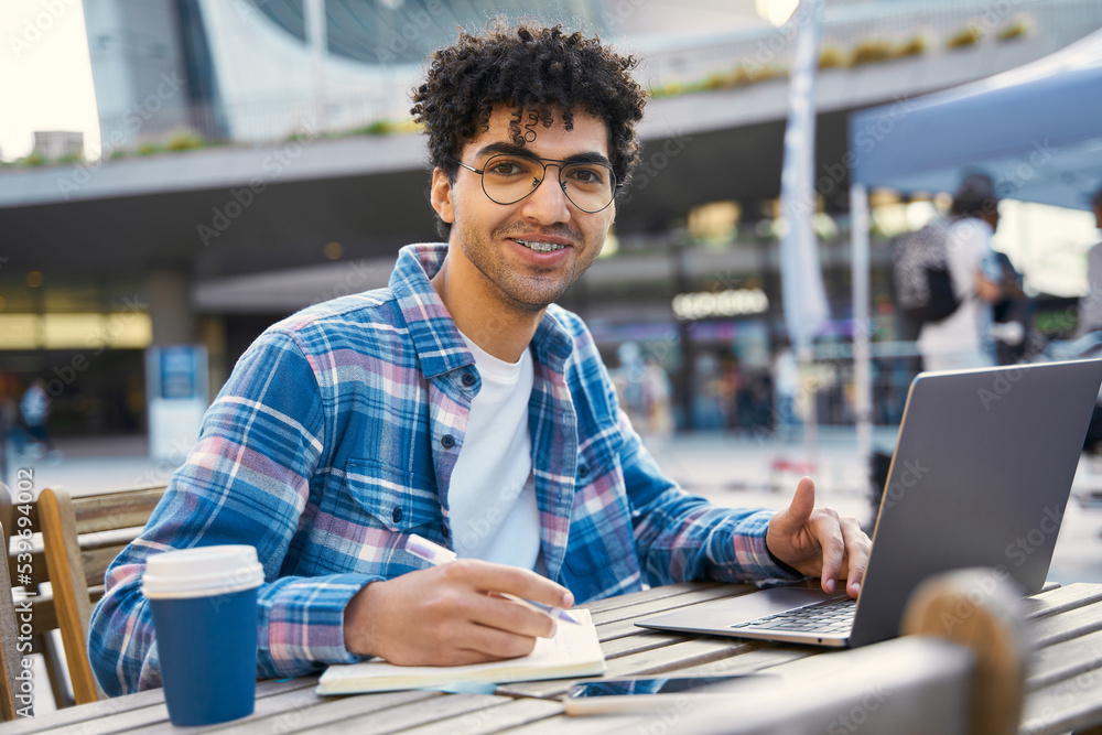 Smiling handsome middle eastern student studying using laptop computer, online education concept ...