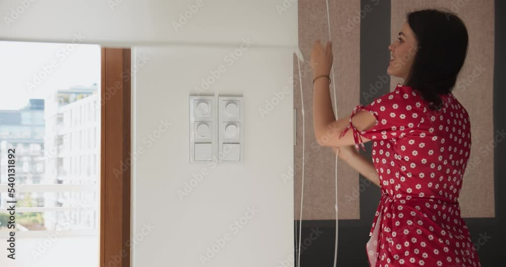 A happy young woman in red dress raises a white roller blinds in the ...