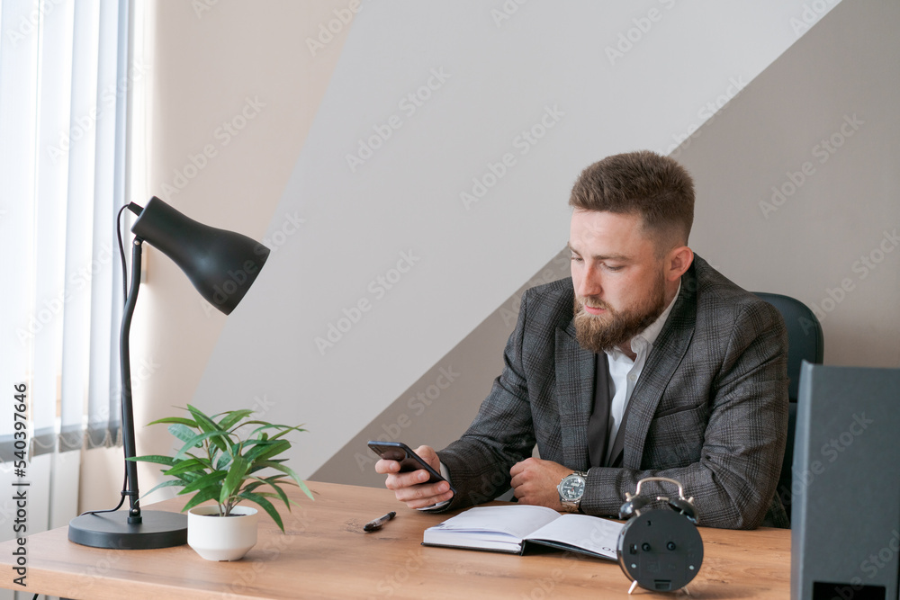 Pensive bearded young man calling by smartphones and writing down notes ...