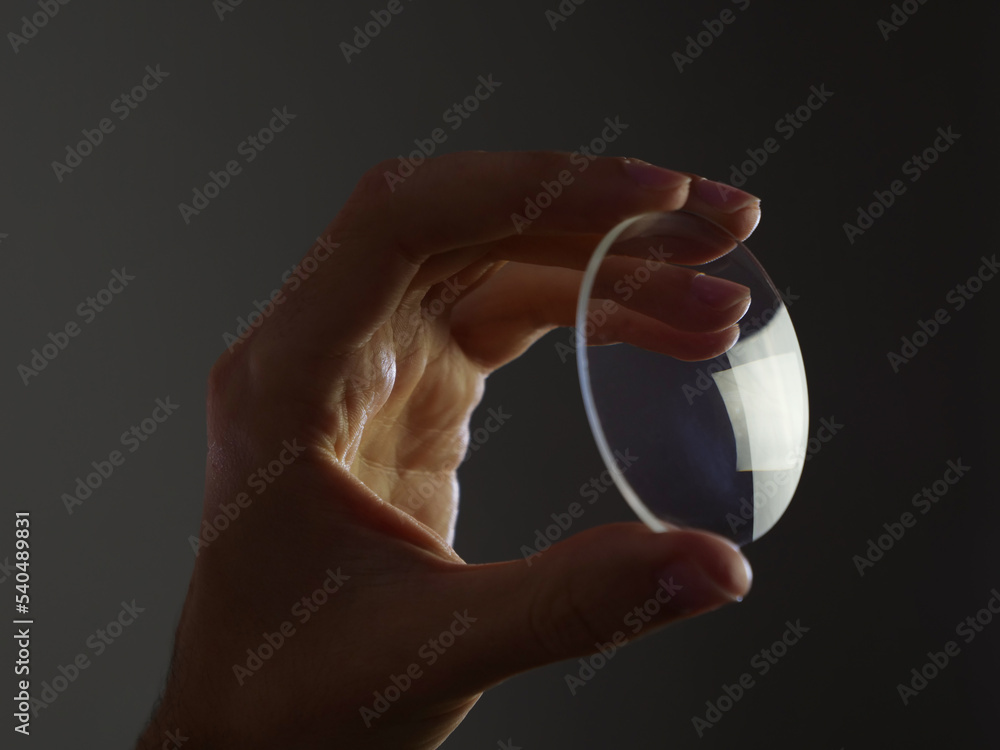 Hand holds an eyewear glass lens, glass prescription lens on dark ...