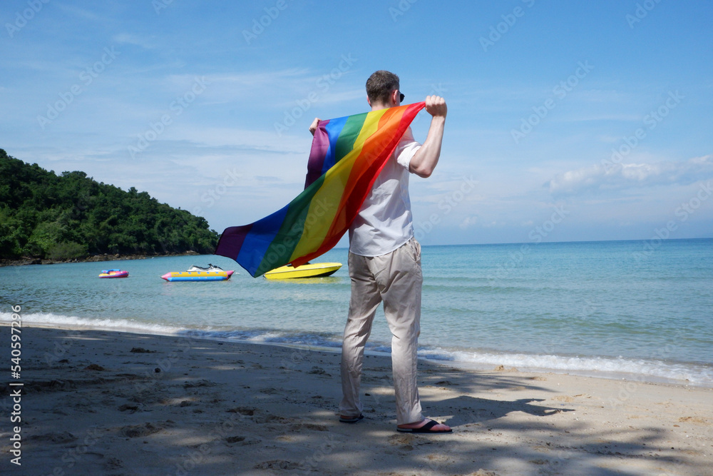 Young man holding a rainbow flag, LGBT symbol, standing on the beach ...