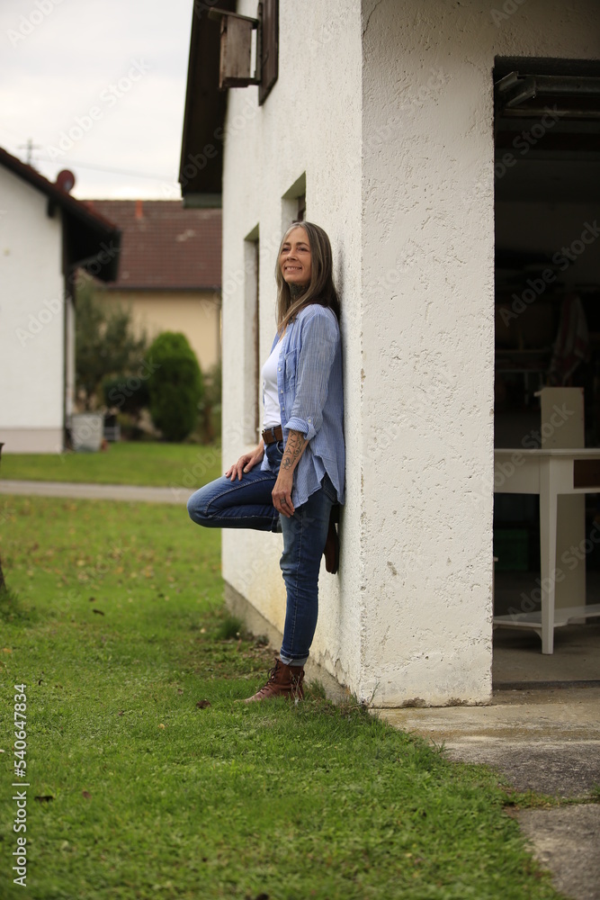 Eine Frau - Ü50 - im besten Alter lehnt an einer Hauswand und sieht in ...