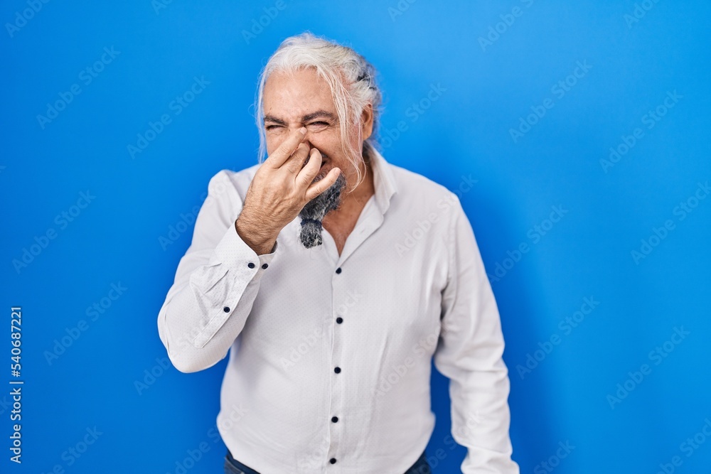 Middle age man with grey hair standing over blue background smelling ...