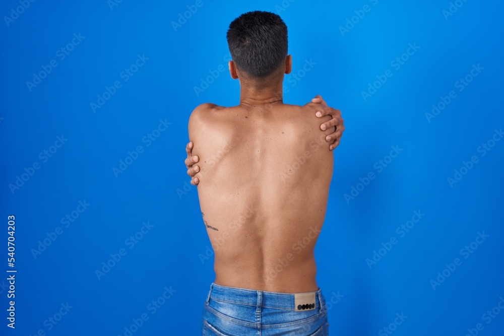 Young hispanic man standing shirtless over blue background hugging ...
