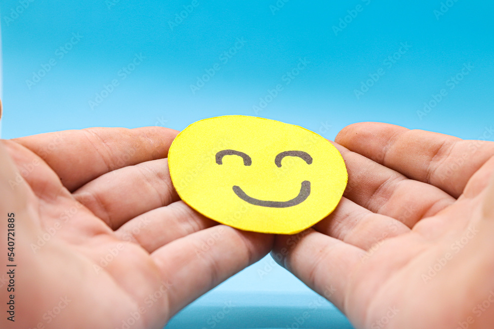Hands holding smiley yellow paper face on a blue background. Happiness ...