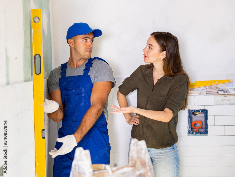 Worker in overall using spirit level, showing flatness of wall to woman ...