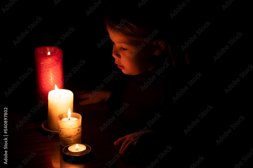 child looks fascinated at the burning flame of a candles in a dark room ...