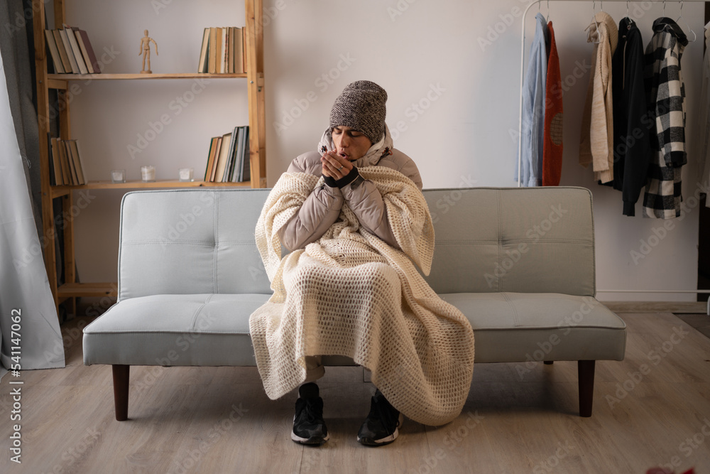 Unwell man in hat and blanket sit in cold living room suffer from heat ...