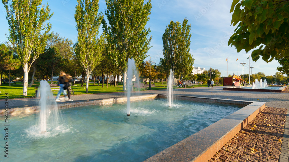 city park, fountain, green lawns and trees, random people walking along ...