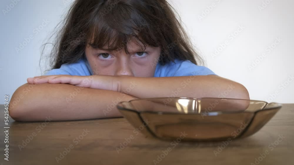 Hungry child with empty plate in room. A hungry upset little girl sits ...