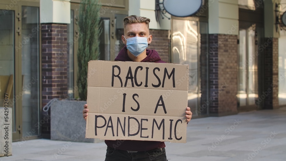 Young man in medical mask stands with a cardboard poster RACISM IS A ...