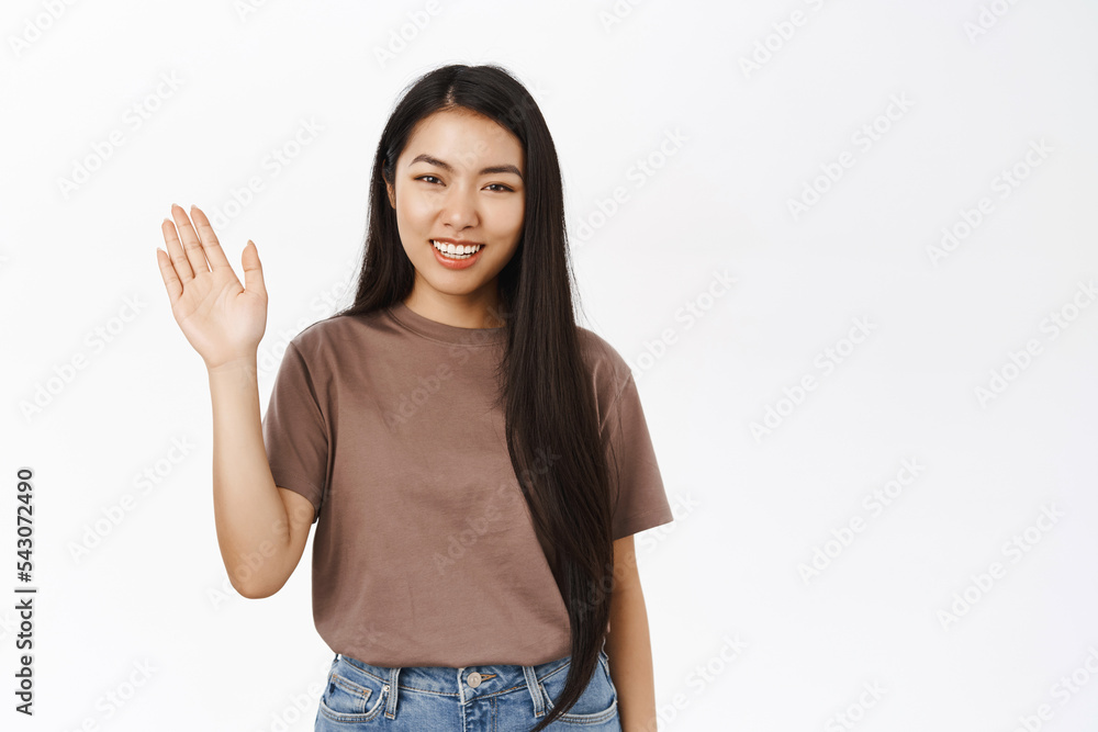 Friendly korean girl waves hand, say hello, hi and smiles at you ...