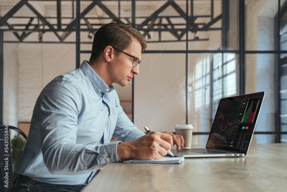 Busy male trader in glasses working online on laptop at his office, financial crypto expert ...