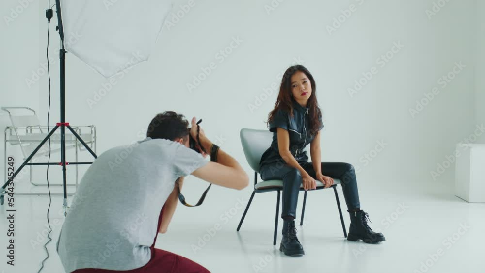 Asian girl sits on chair, looks to photographer with camera. Studio ...