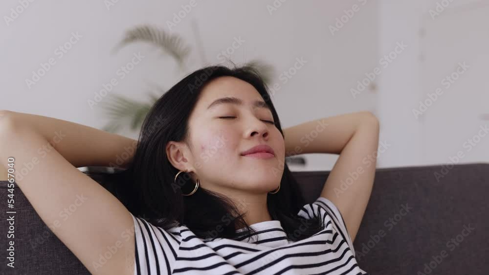 Young chinesechie woman with hands behind head relaxing on sofa at home ...