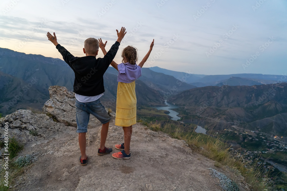 Happy boy and girl hikers standing with raised hands on rock and enjoying view of nature ...