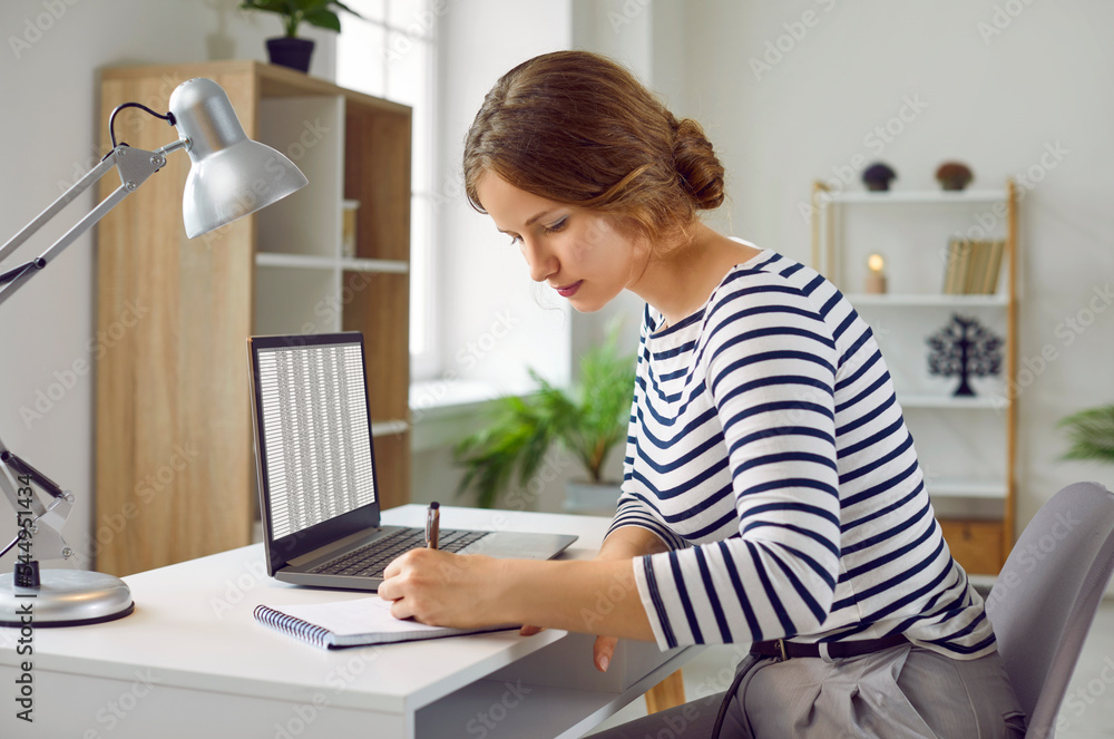 Left-handed woman makes notes in her notebook while working on laptop ...