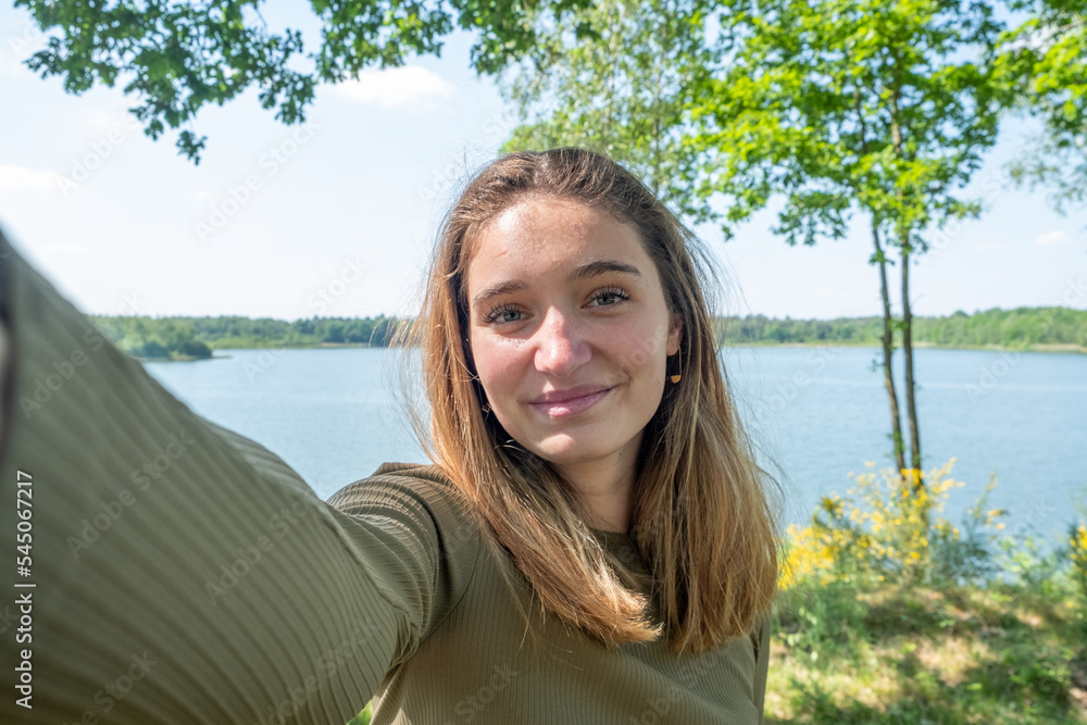 Beautiful young brunette Caucasian woman taking a selfie with smartphone outdoors on a forest ...