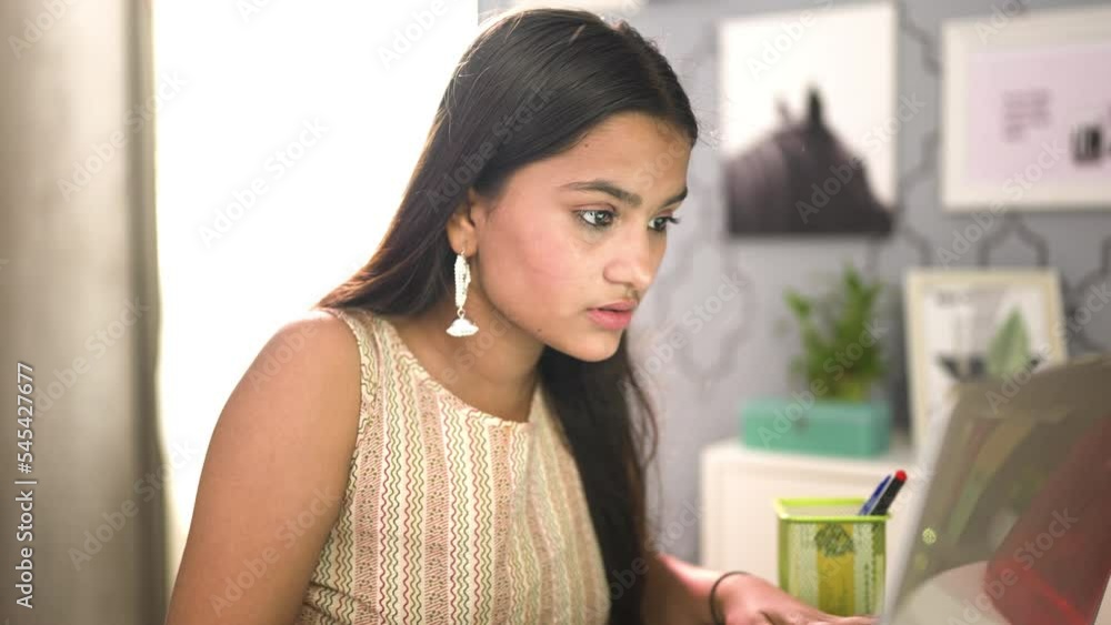 Beautiful young Indian girl at home studying, typing on laptop ...