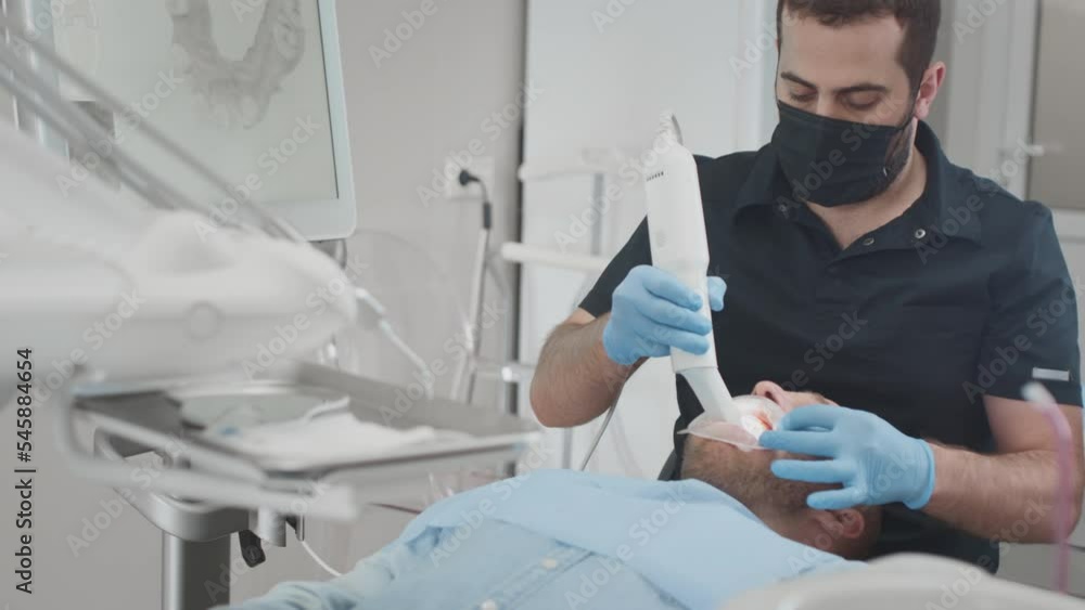 A male orthodontist doctor scans a man's teeth and jaw in the clinic's ...