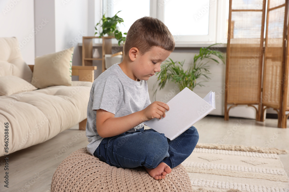 Boy with poor posture reading book on beige pouf in living room ...