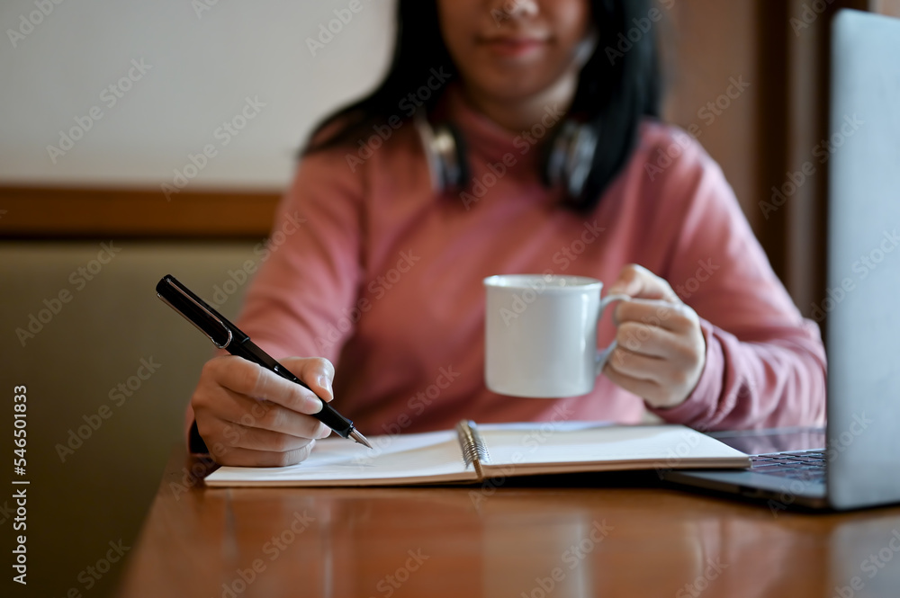Young Asian female taking notes on her notebook while sipping coffee ...