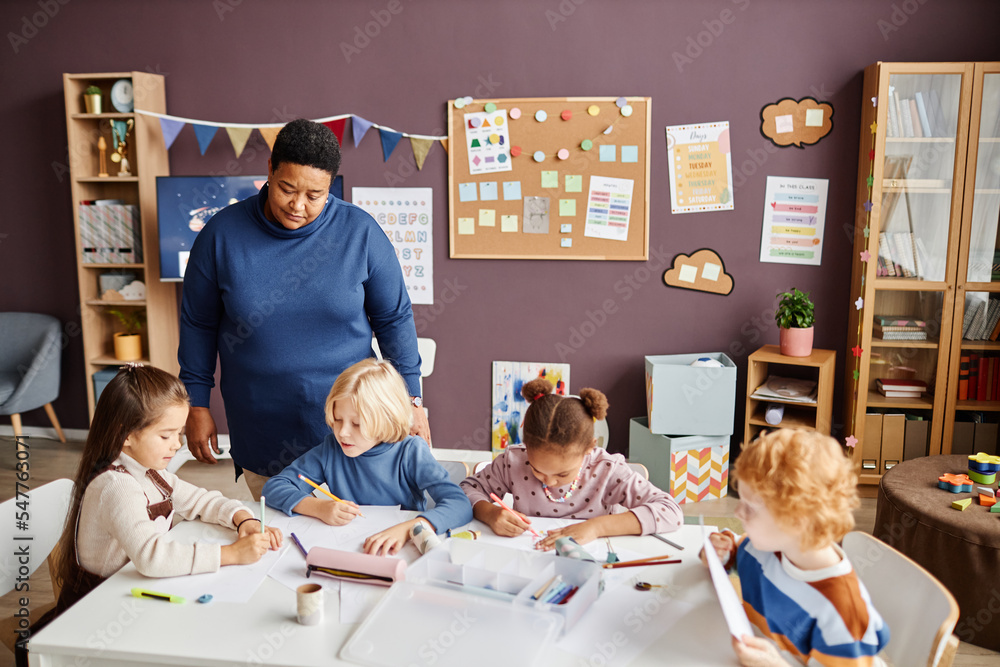 Mature African American nursery teacher standing by table where group ...
