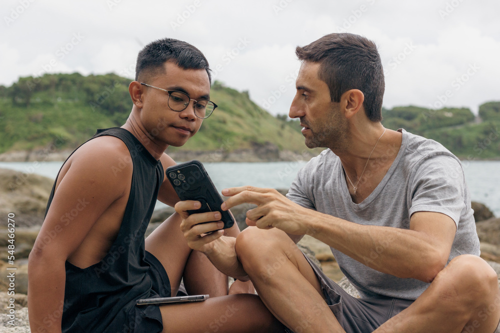 Young man showing cellphone to Filipino friend sitting on rocks by the ...
