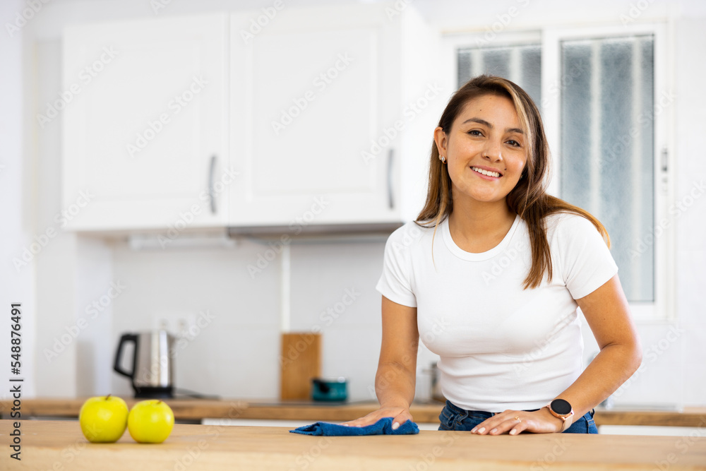 Portrait of cheerful young Latina cleaning table with cloth and spray ...