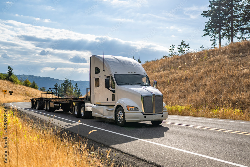 Big rig white semi truck transporting two empty flat bed semi trailer ...