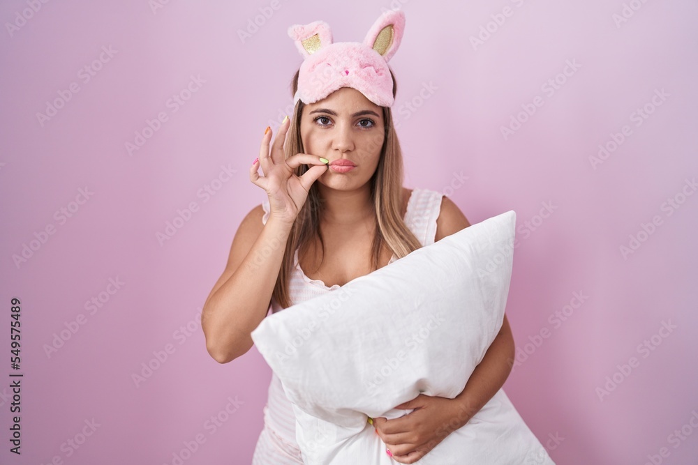 Young blonde woman wearing pyjama hugging pillow mouth and lips shut as ...