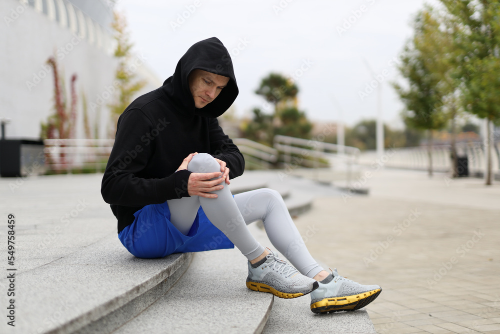 Handsome muscular male jogger wearing warm training outfit touching his knee in pain with ...