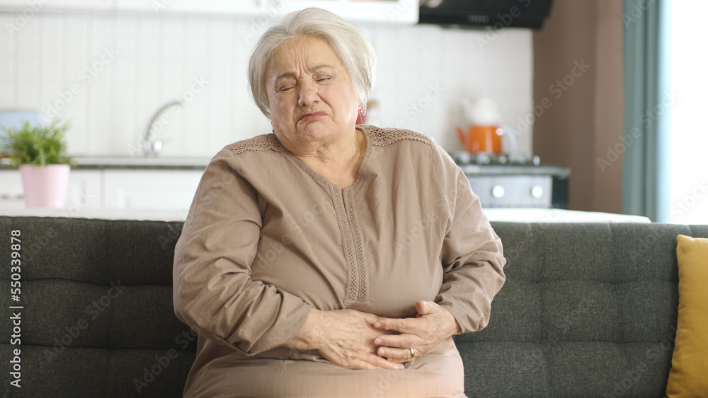 Lonely old woman sitting alone on sofa at home having digestive system ...