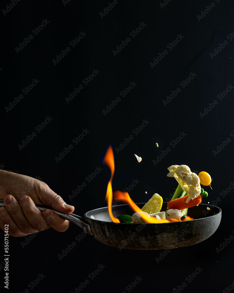 Tossing vegetables in a cooking pan with flames of fire around them ...