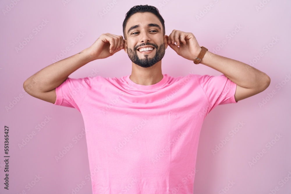 Hispanic young man standing over pink background smiling pulling ears ...