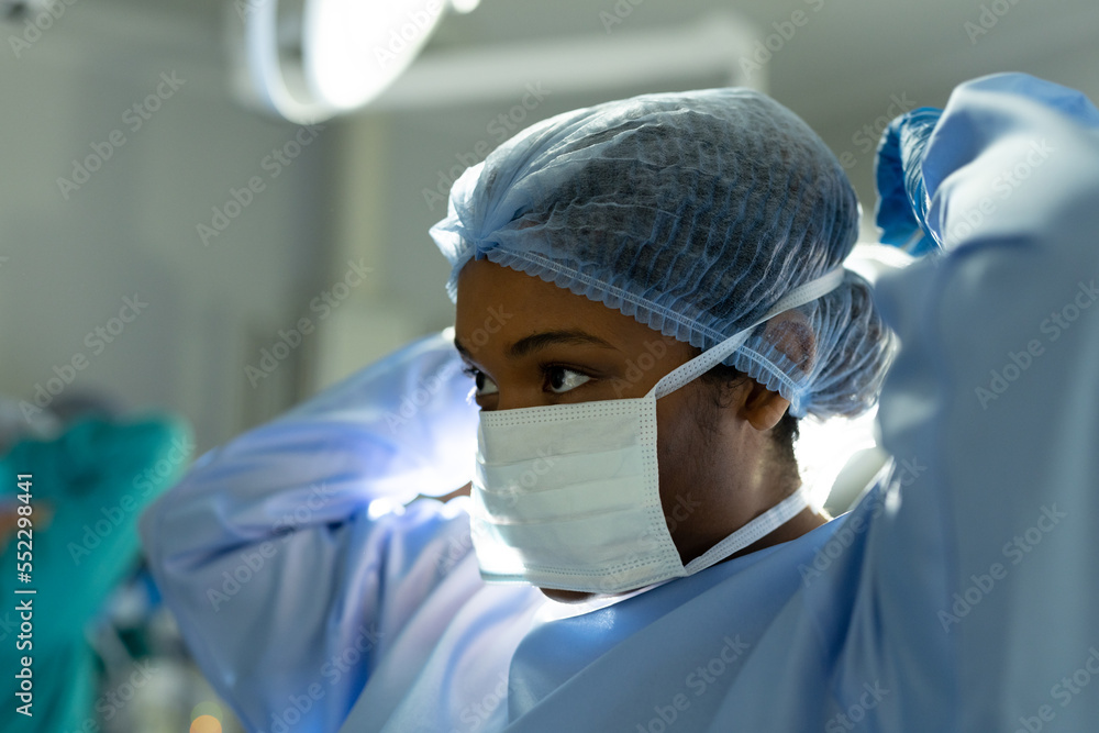 Biracial female surgeon in surgical cap and gown putting on mask in ...