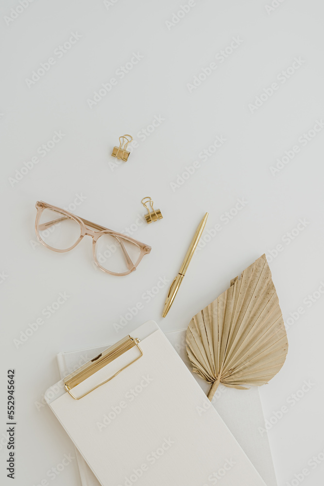 Flatlay of clipboard with blank copy space paper sheet. Tan fan leaf, glasses, pen, clips on ...
