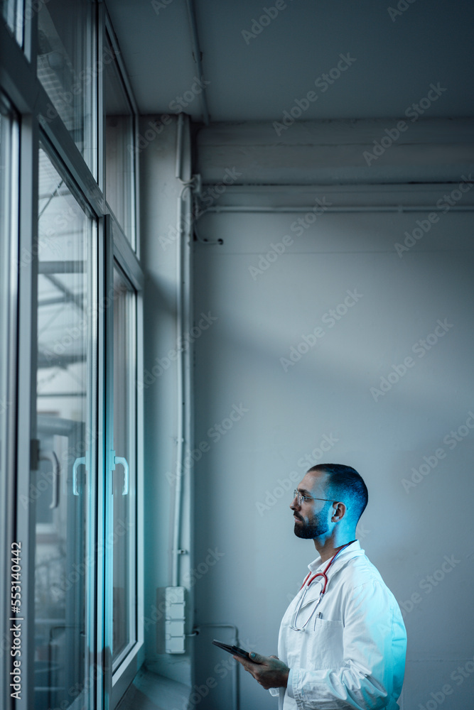 Thoughtful doctor holding tablet PC looking through window in front of ...