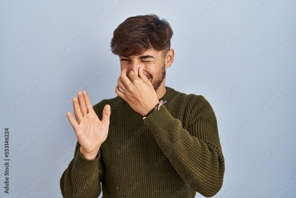 Arab man with beard standing over blue background smelling something ...