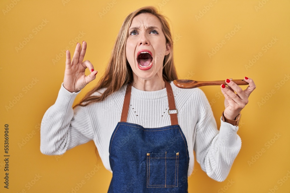 Young blonde woman wearing apron tasting food holding wooden spoon ...